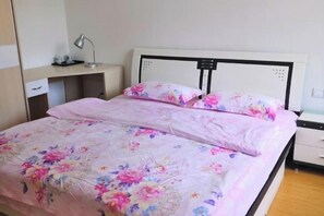 Cama bem arrumada com roupa de cama florida, mesa de cabeceira com abajur e gaveteiro pequeno.