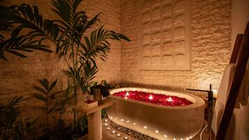 Espaço de spa com parede de pedra, banheira cheia de pétalas de rosa e uma planta grande.