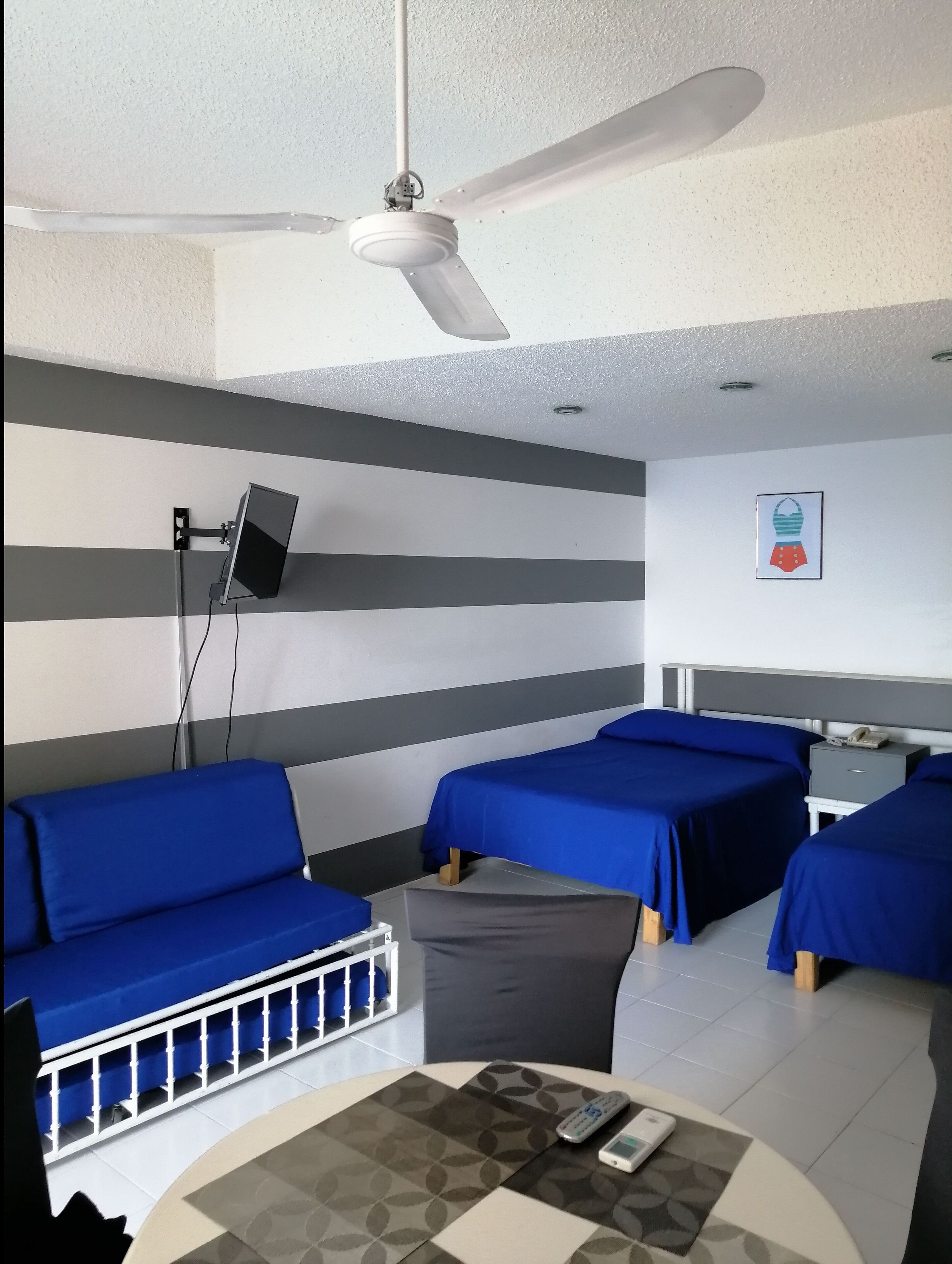 Quarto de hotel com duas camas, um sofá, uma TV fixada na parede, um ventilador de teto e uma pequena mesa com um controle remoto.