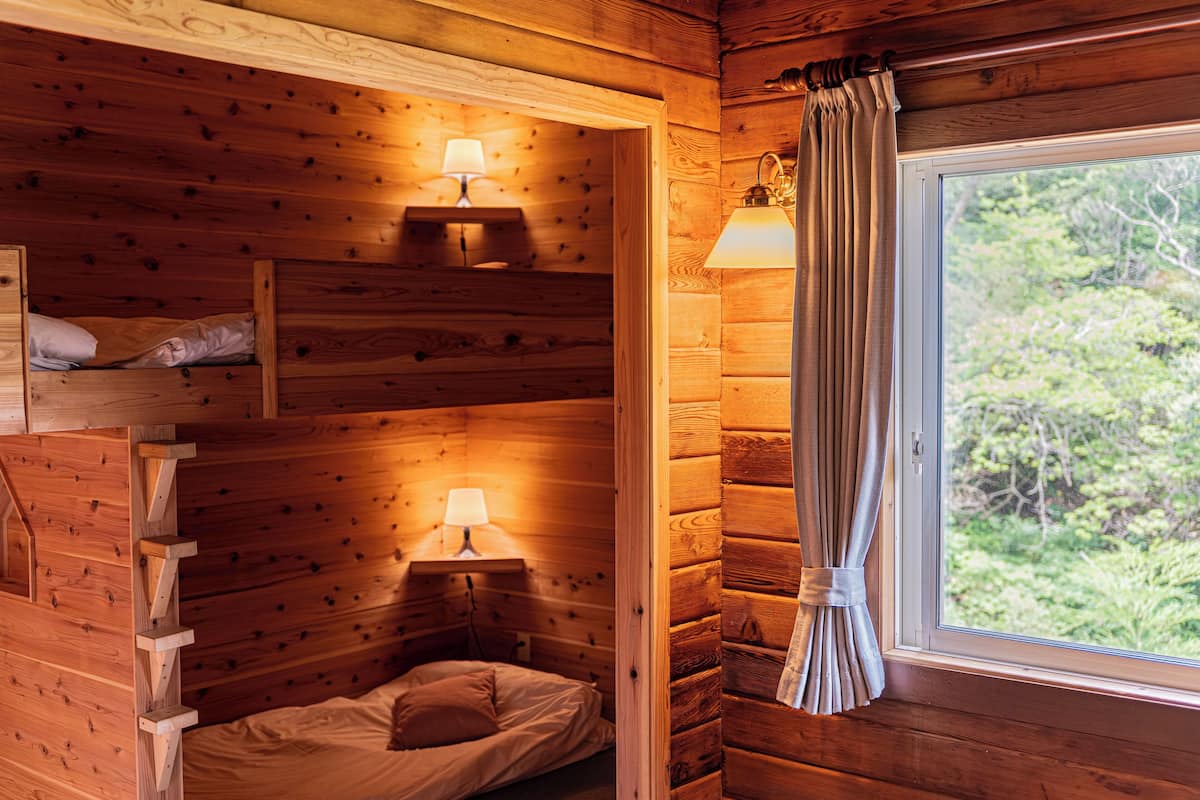 Quarto de cabana de madeira com cama, mesa de cabeceira com abajur e janela com cortinas.