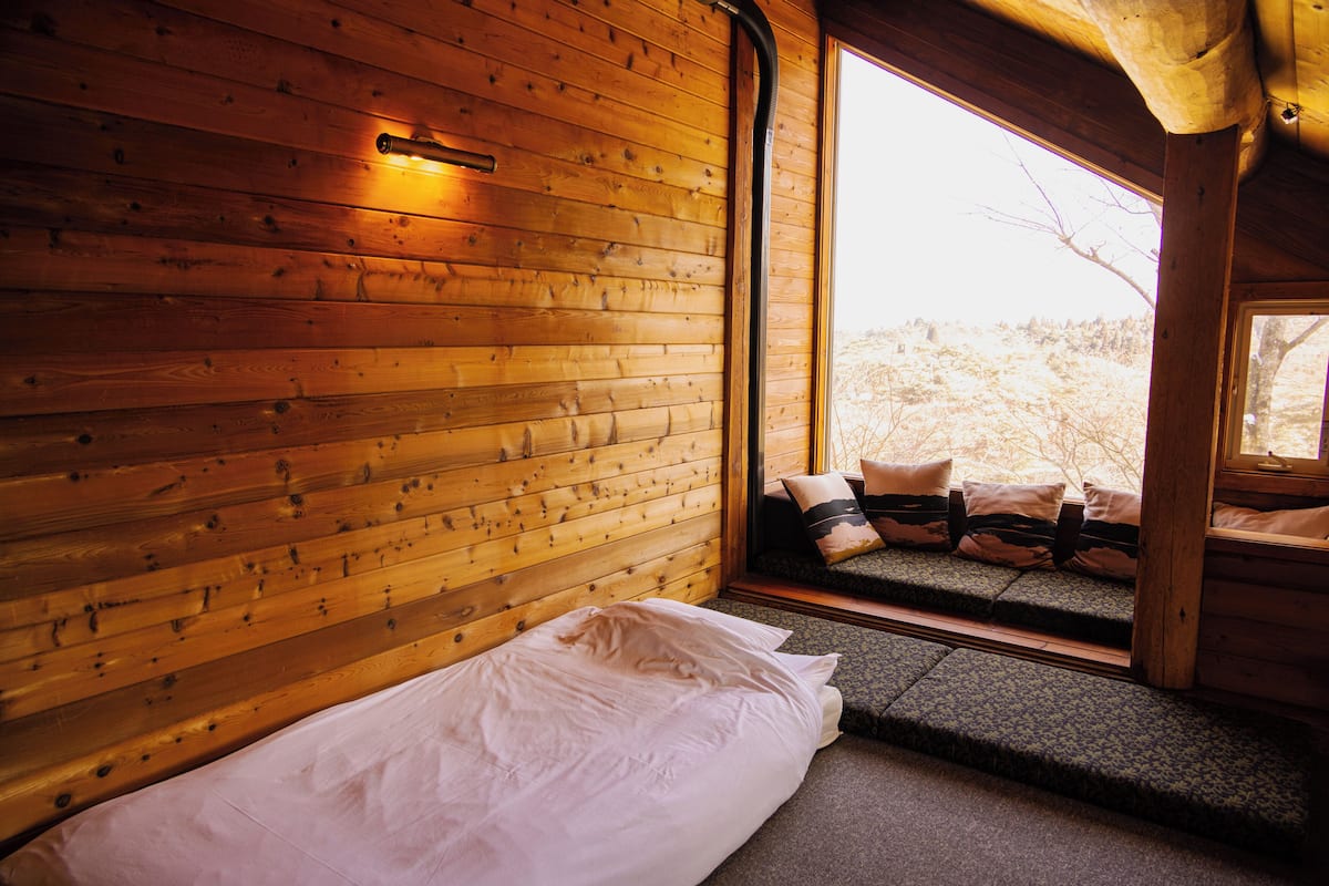 Quarto de cabana de madeira com uma cama, janela com vista para árvores e uma pequena mesa com cadeiras.