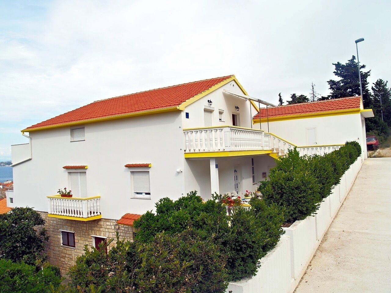 Um edifício de dois andares com telhado de telhas vermelhas, paredes brancas e detalhes amarelos. Possui uma varanda com balaústres brancos e uma pequena área de jardim com arbustos verdes.
