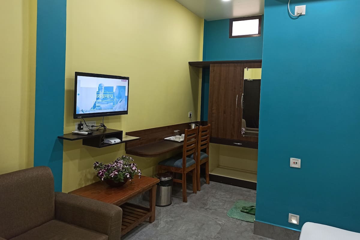 Um quarto pequeno com uma escrivaninha, televisão, cadeira, sofá, lixeira e janela.