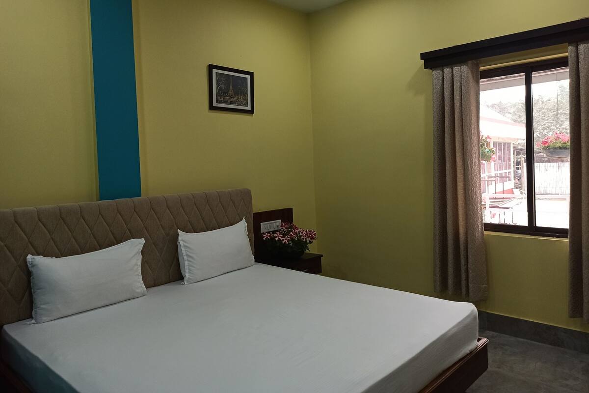Quarto com cama, mesa de cabeceira com um vaso de flores, quadro na parede e janela com cortinas.