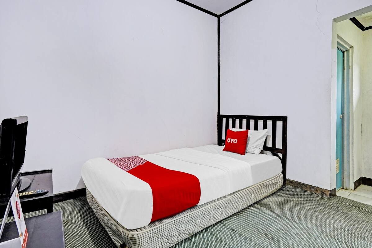 Uma cama de solteiro com colcha branca e vermelha, cabeceira preta e travesseiro vermelho com a palavra "OYO".