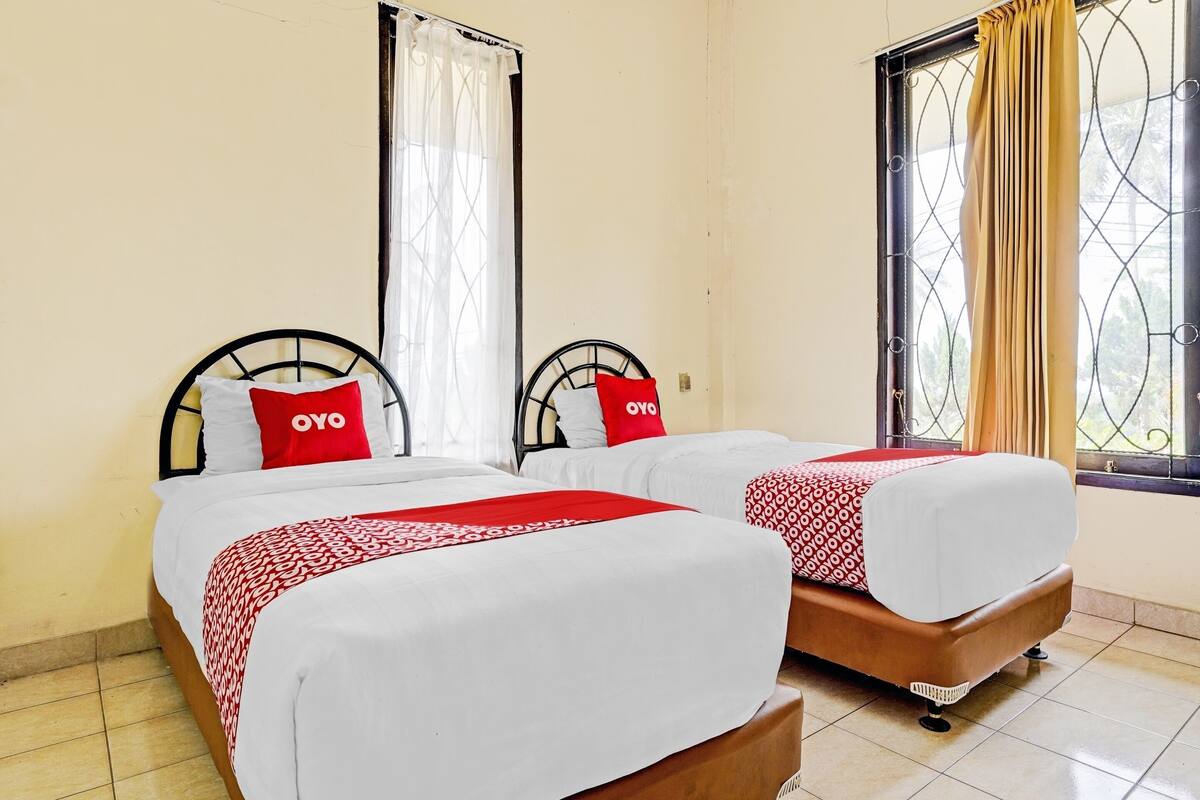 Duas camas de solteiro com estrutura de metal, roupa de cama branca, almofadas vermelhas com a marca 'OYO' e uma janela com grade de metal e cortinas.