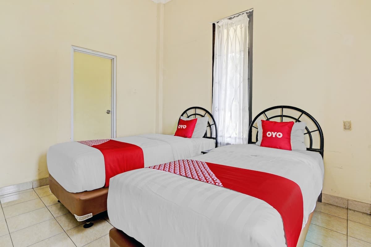 Quarto de hotel com duas camas de solteiro, cada uma com colcha e almofadas vermelhas e brancas. Há uma porta e uma janela com cortinas.