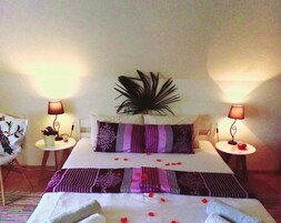 Quarto de hotel com cama bem arrumada, duas luminárias de cabeceira, uma planta decorativa e uma mesinha com frutas.
