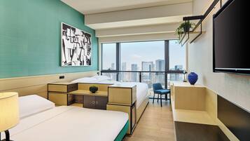 Quarto de hotel com duas camas, uma janela ampla com vista para a cidade, uma televisão montada e uma pequena escrivaninha com uma planta.