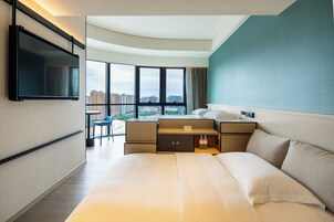 Quarto de hotel moderno com uma cama grande, televisão fixada na parede, mesa com cadeira e vista da cidade através de amplas janelas.