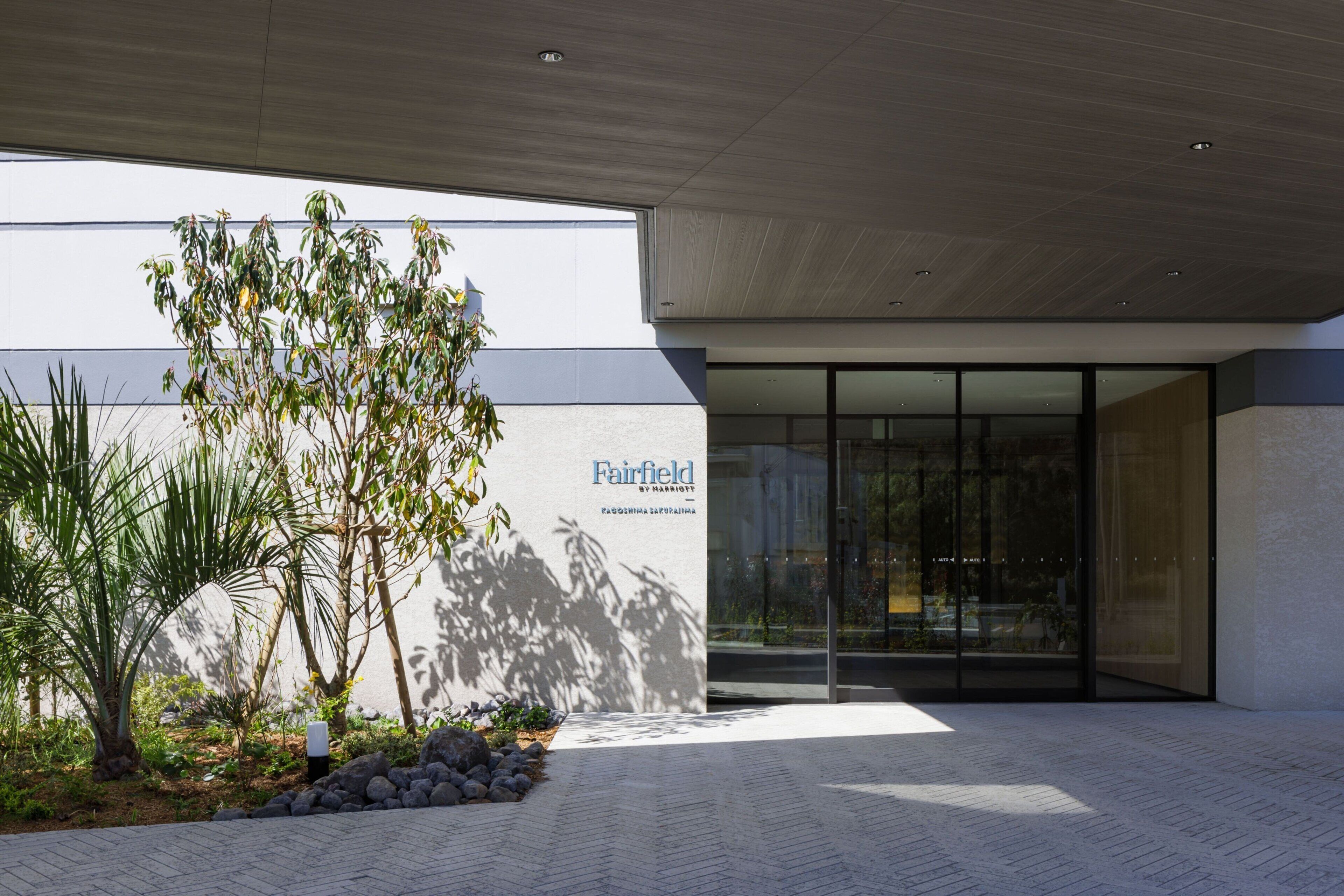 Entrada de um edifício moderno com uma placa indicando "Fairfield by Marriott", área ajardinada com plantas e pedras, e um caminho pavimentado que leva à entrada.