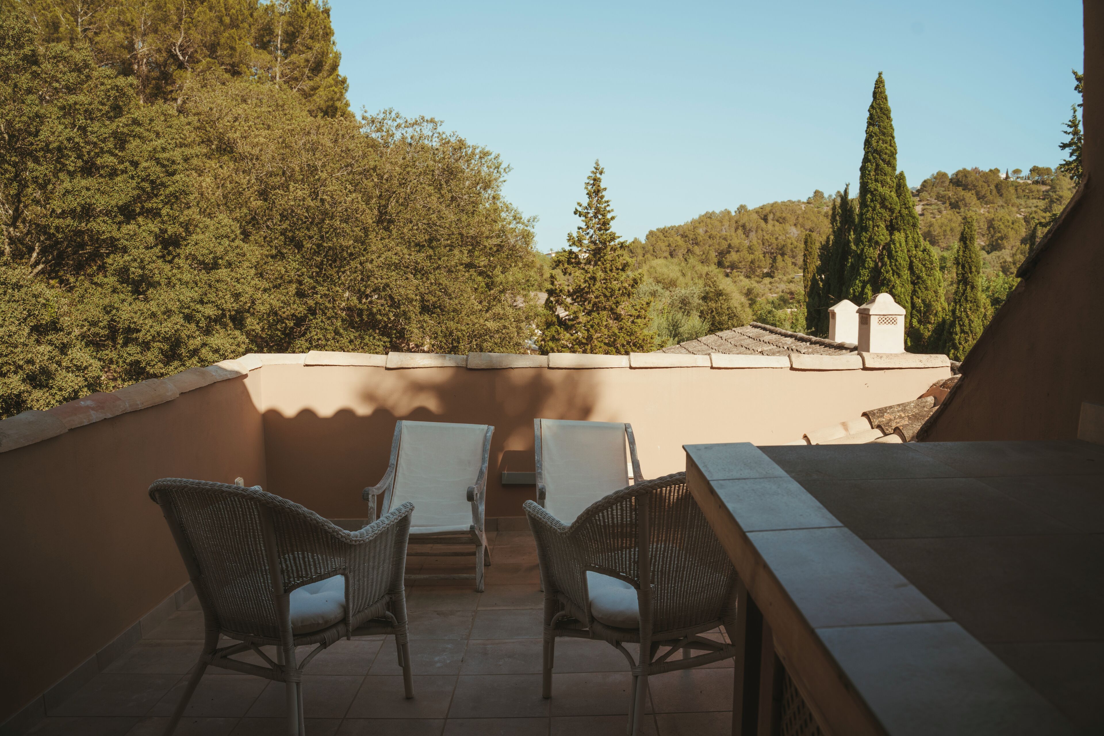 Terraço com cadeiras de vime e uma mesa, com vista para uma área arborizada sob um céu claro.