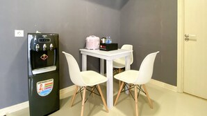 Quarto compacto com geladeira preta, mesa branca e duas cadeiras brancas, porta fechada.