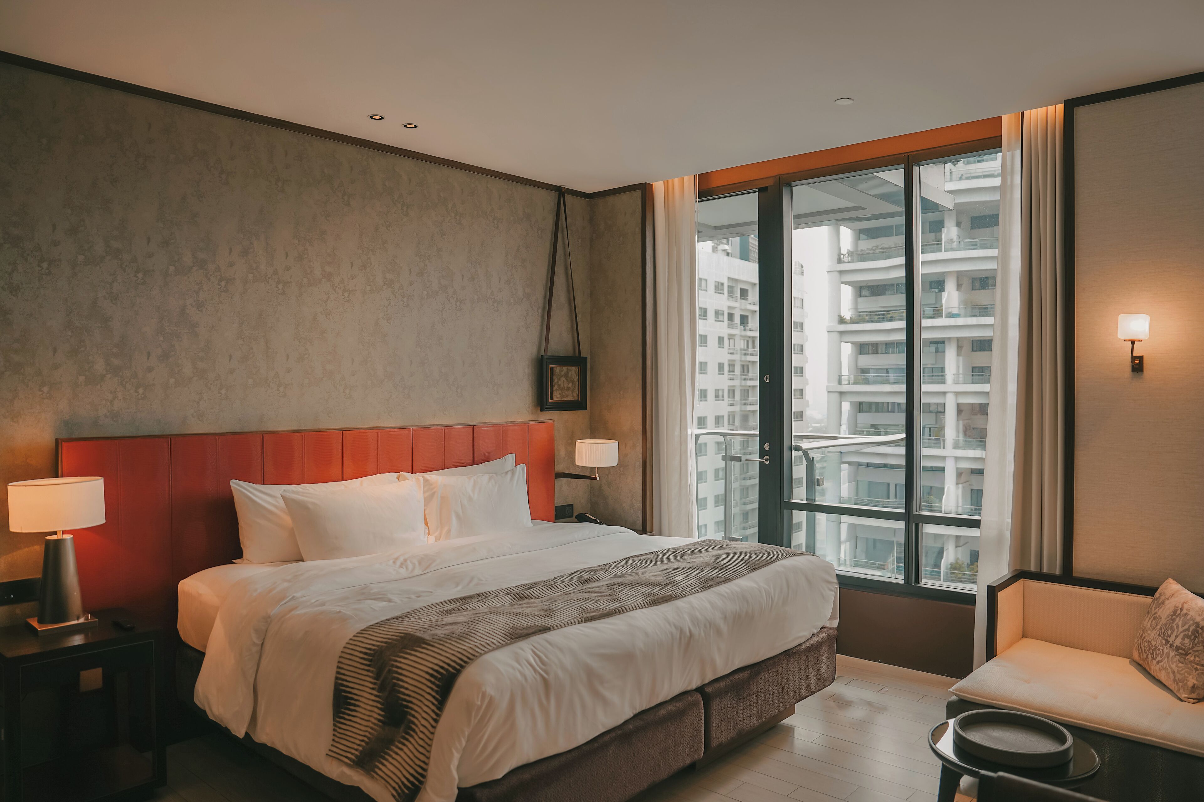 Quarto de hotel com uma cama grande, uma mesa de cabeceira com abajur, um sofá e vista para a cidade através de uma janela ampla.