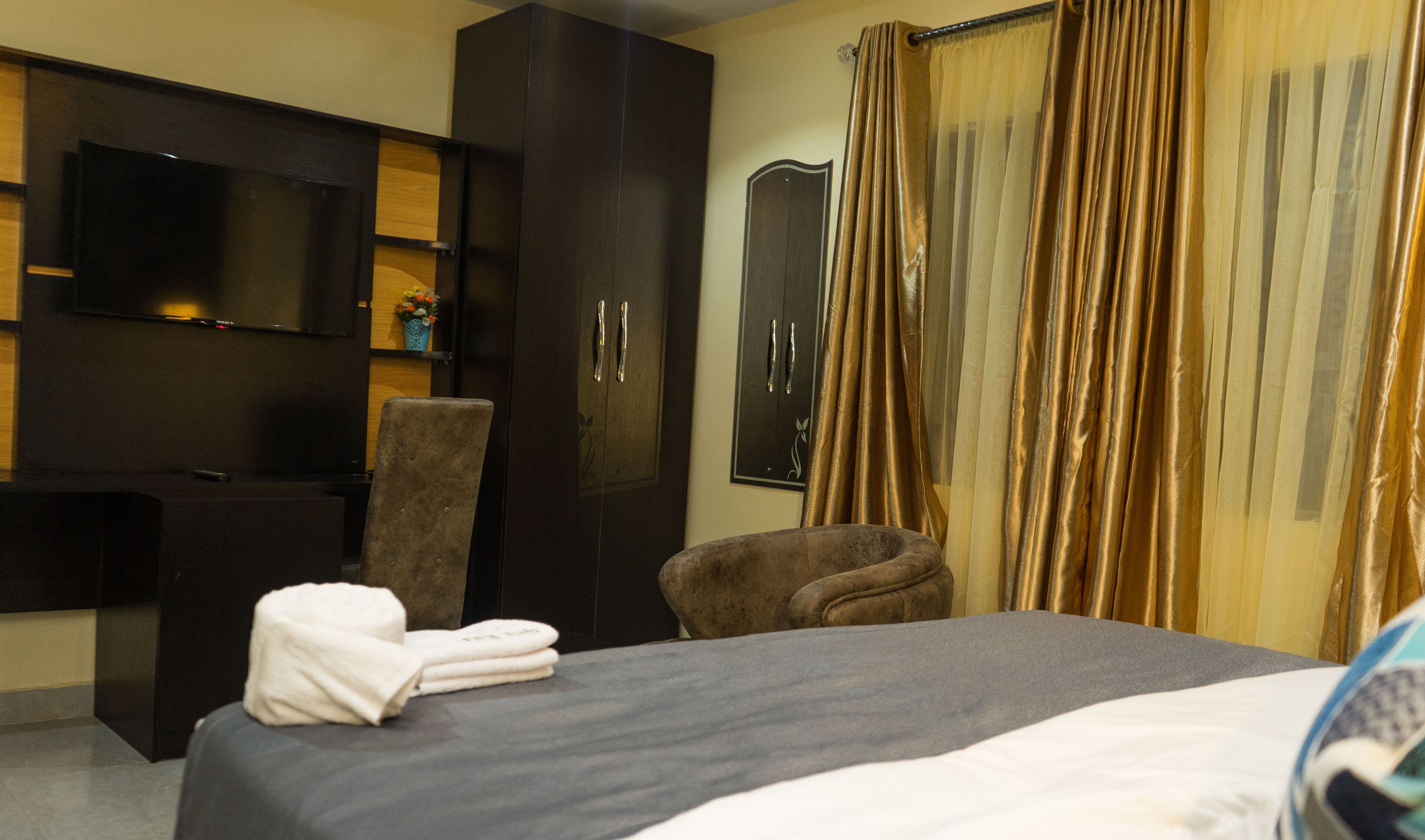 Quarto de hotel com cama, televisão na parede, guarda-roupa, cadeira e cortina.