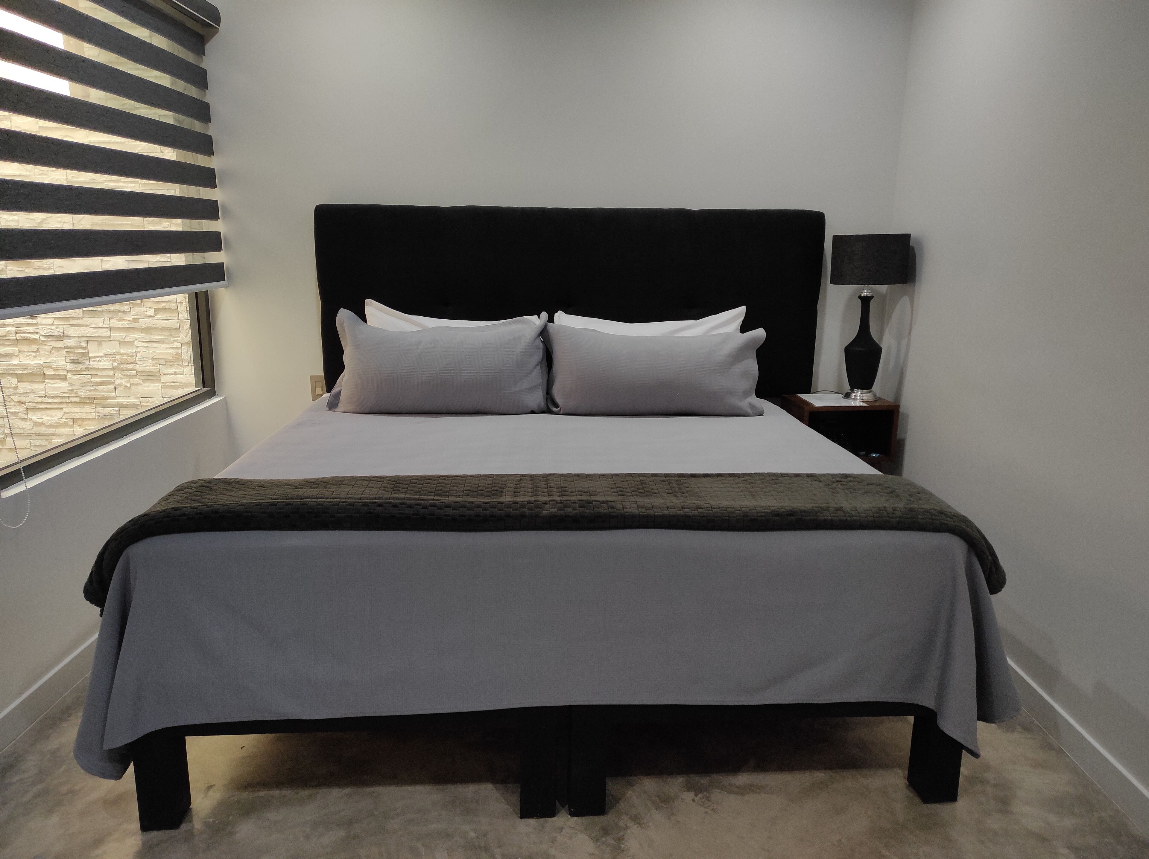 Cama bem arrumada com cabeceira escura, almofadas brancas e manta cinza. Tem uma mesa de cabeceira com luminária e uma pequena estante. Uma janela com persianas é visível ao fundo.