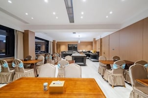 Uma sala de jantar de hotel com várias mesas e cadeiras, uma cozinha ao fundo e uma janela grande à esquerda.