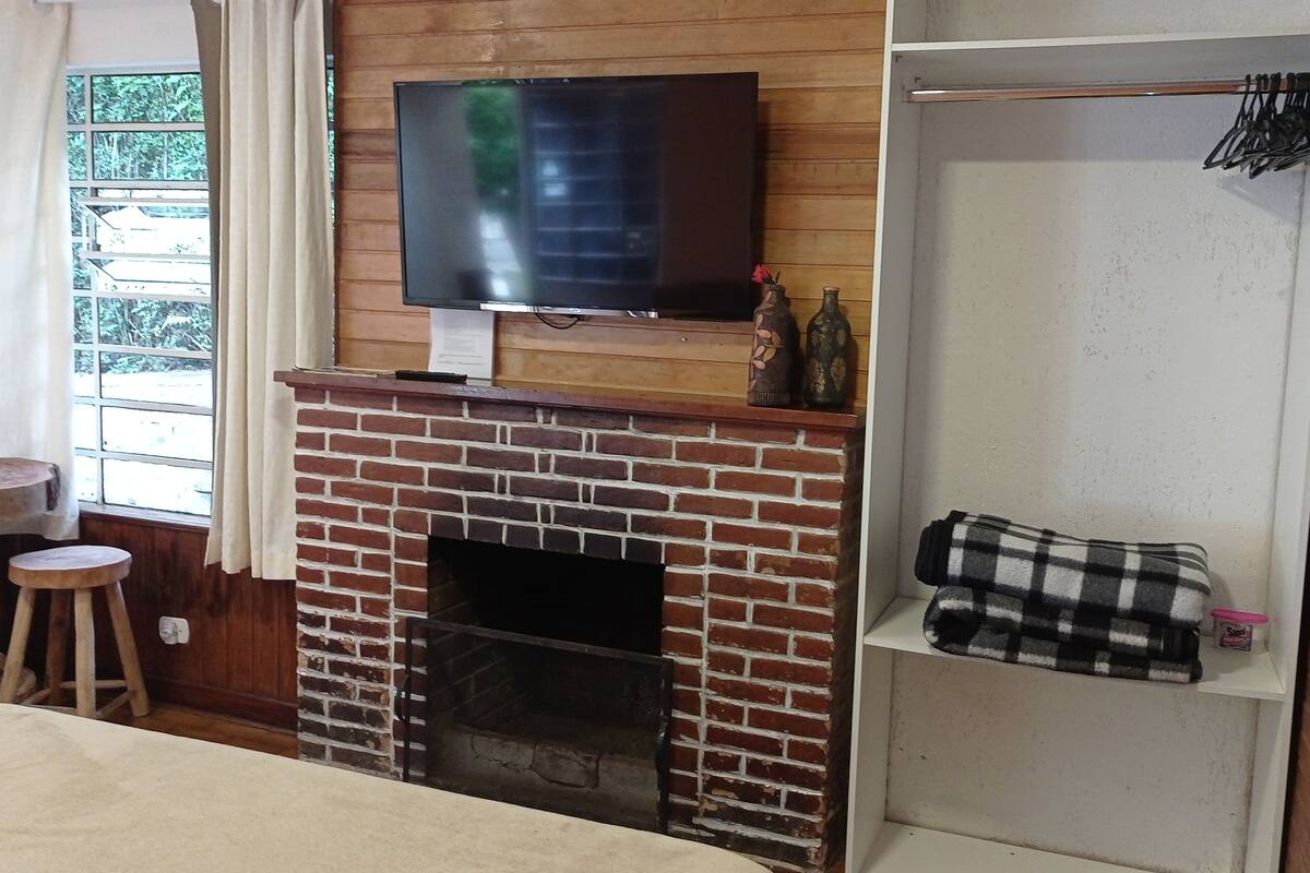 Quarto com lareira de tijolos, televisão na parede, janela com cortinas e um banquinho de madeira.