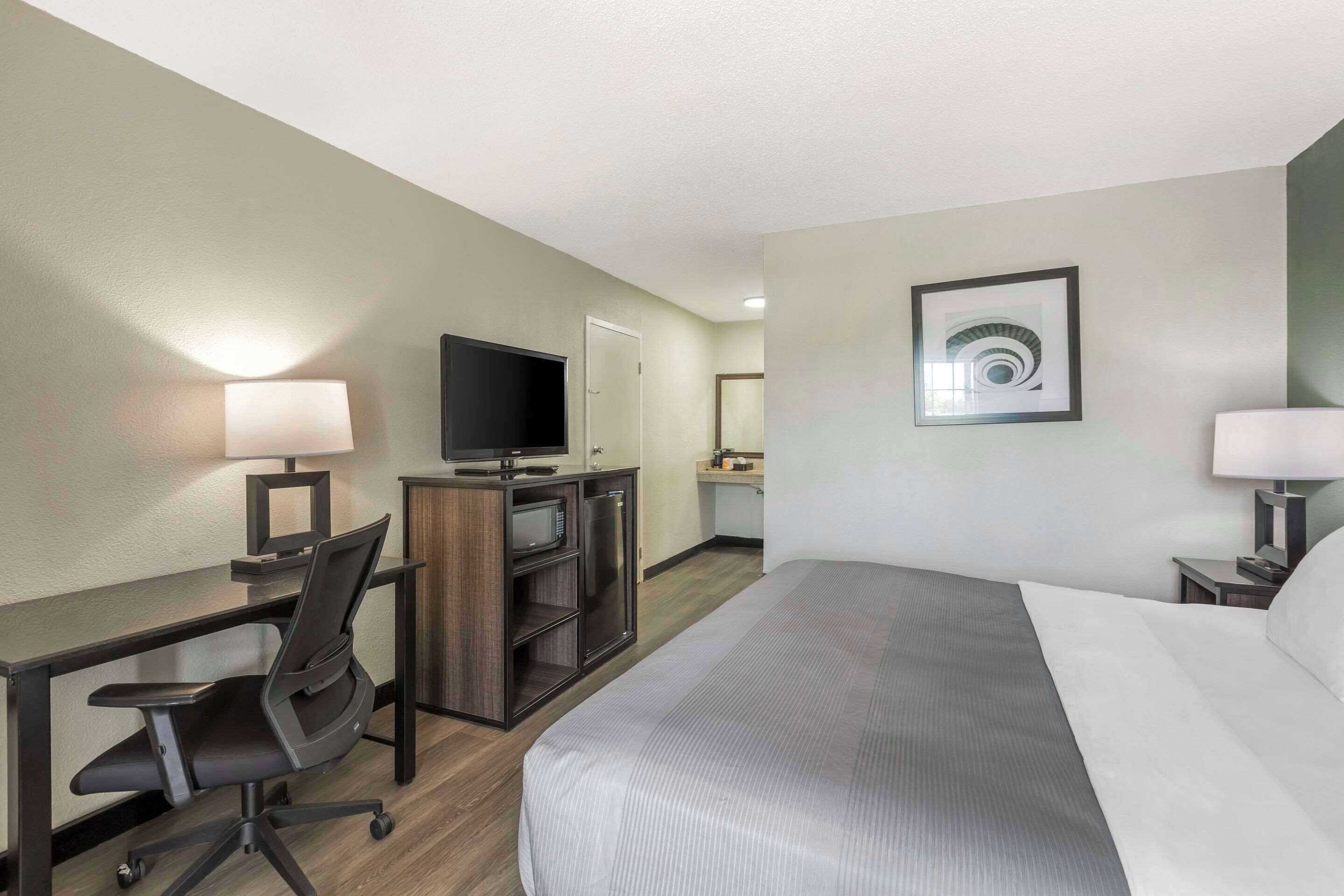 Quarto de hotel com cama, escrivaninha, cadeira, televisão e uma pequena cozinha compacta.