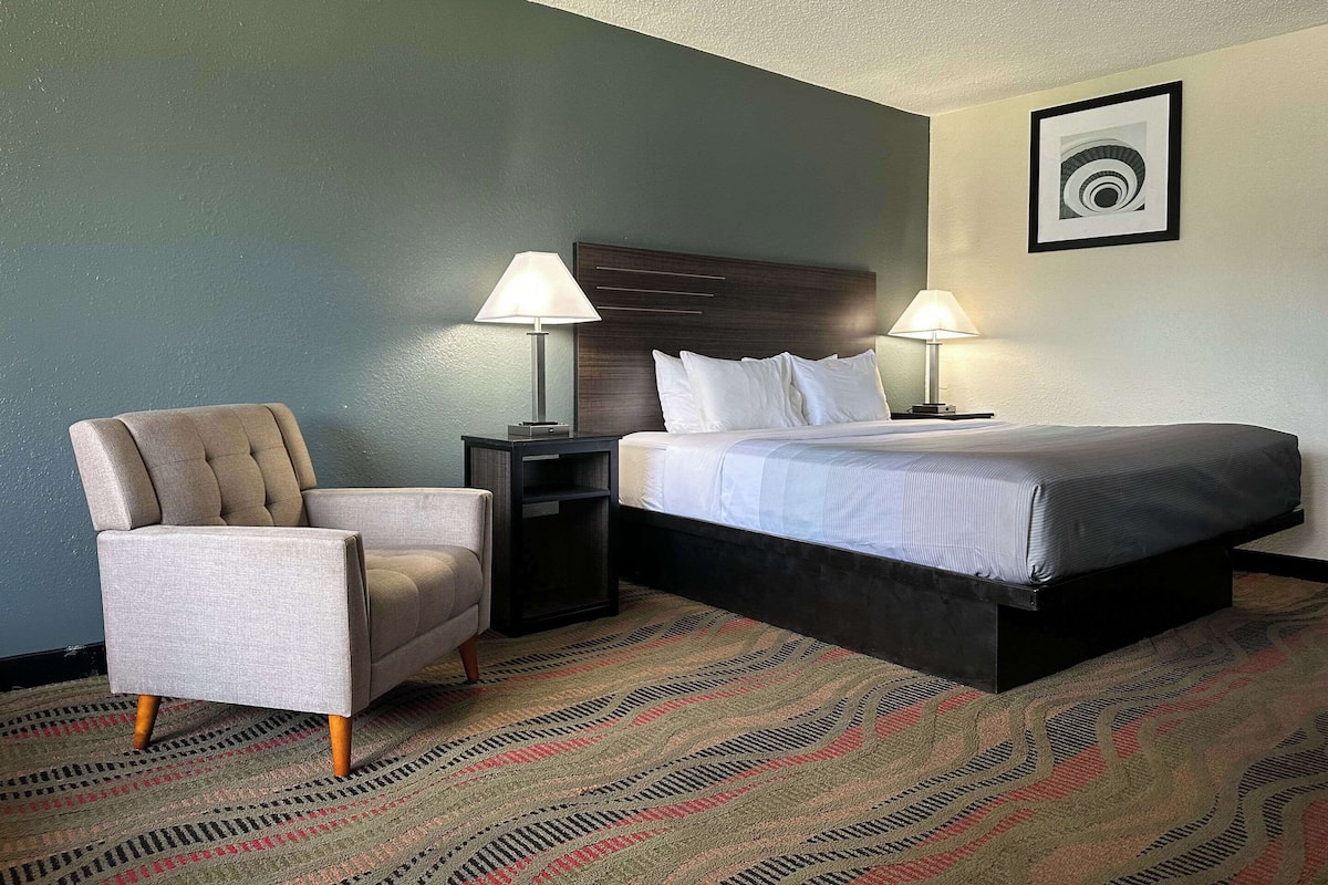 Quarto de hotel com uma cama, duas mesas de cabeceira com luminárias, uma poltrona e um tapete estampado.