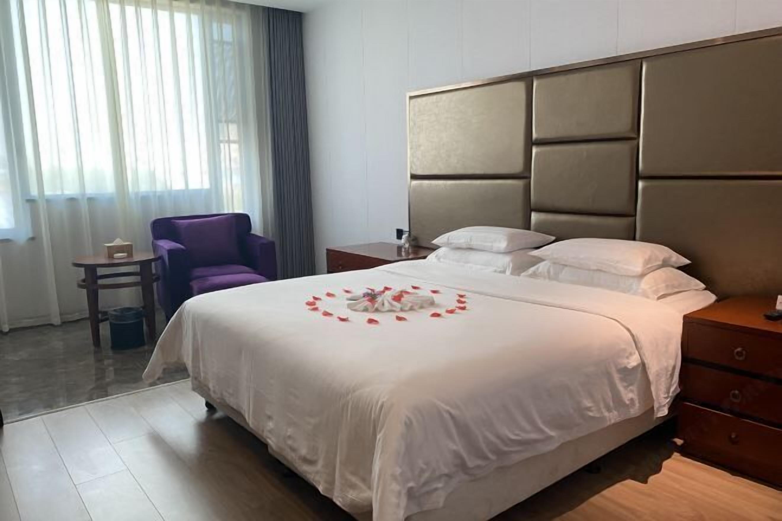 Quarto de hotel com uma cama grande, uma mesa de cabeceira de madeira, uma poltrona roxa e uma pequena mesa com abajur.