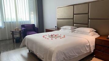 Quarto de hotel com uma cama grande, uma mesa de cabeceira de madeira, uma poltrona roxa e uma pequena mesa com abajur.