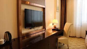 Quarto de hotel com televisão de tela plana presa a um armário de madeira, uma escrivaninha com abajur, uma cadeira e uma cama com carpete estampado.