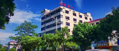 Um edifício com vários andares com uma placa vermelha no topo, cercado por árvores e veículos na rua.