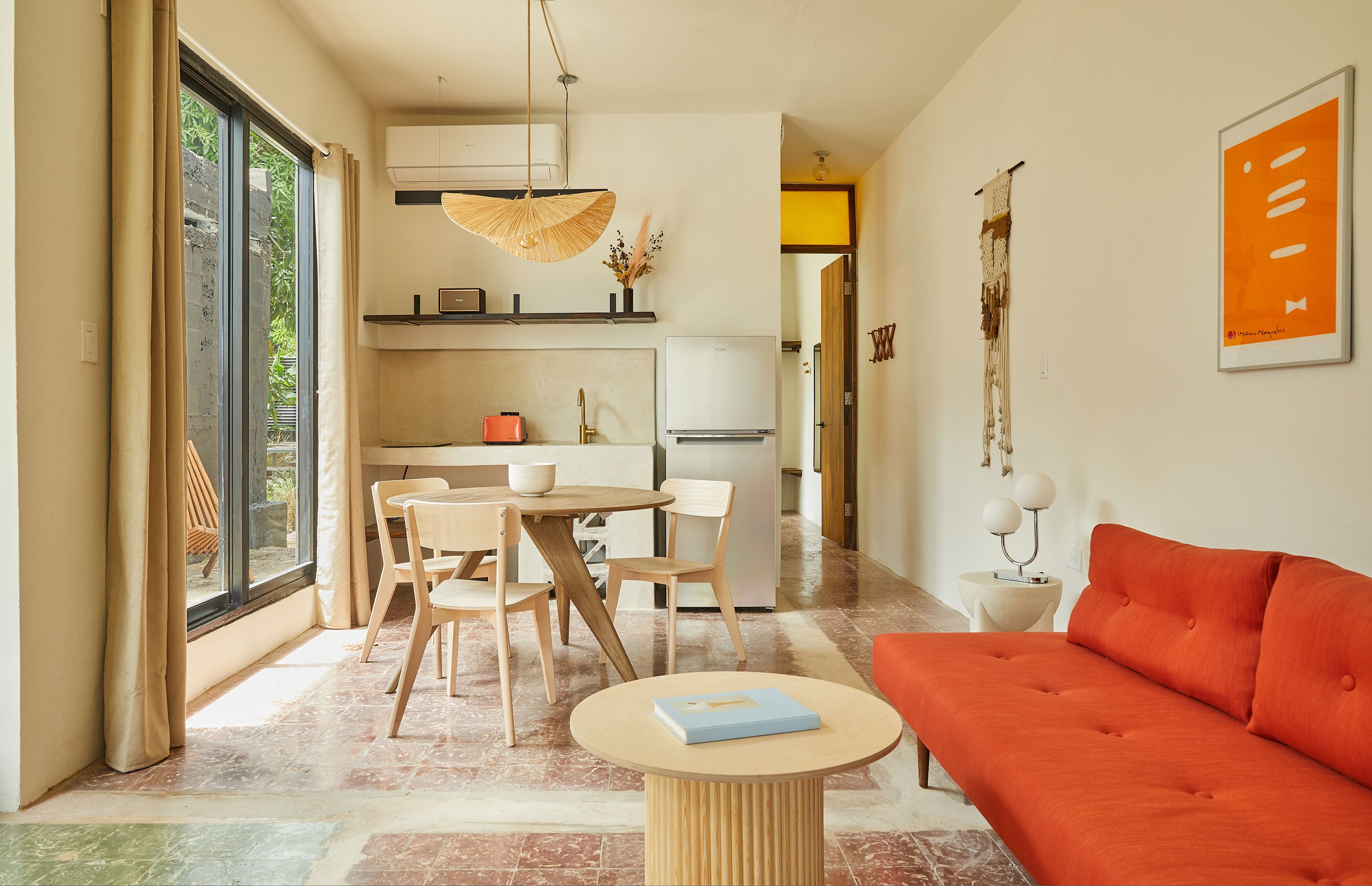 Uma sala de estar moderna com um sofá vermelho, uma área de jantar com mesa e cadeiras, e uma cozinha com geladeira e luminária pendente.