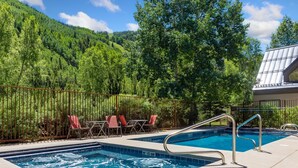 Uma piscina com uma escada de metal, rodeada por cadeiras e um cercado, com uma encosta arborizada ao fundo.