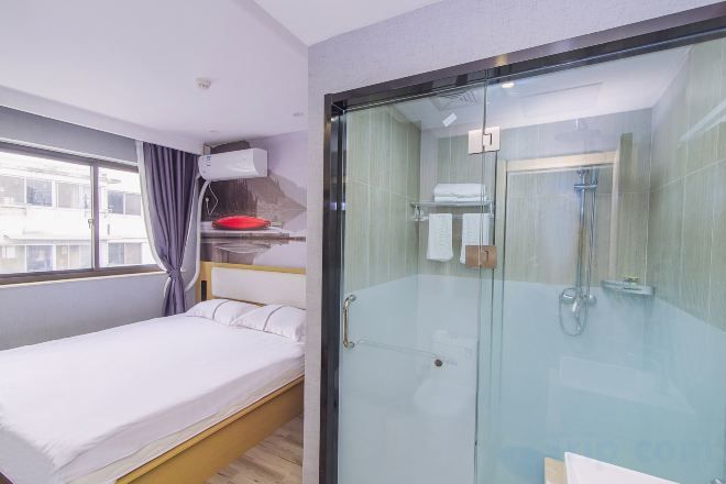 Quarto de hotel com cama, chuveiro com proteção de vidro e janela com cortinas.