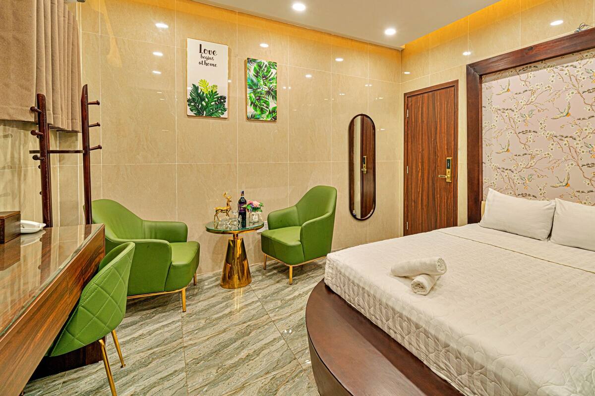 Quarto de hotel com uma cama grande, duas cadeiras verdes, uma mesa redonda pequena com um objeto decorativo e uma porta de madeira.