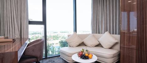 Quarto de hotel moderno com uma janela grande, um sofá, uma mesa pequena com frutas e uma escrivaninha.