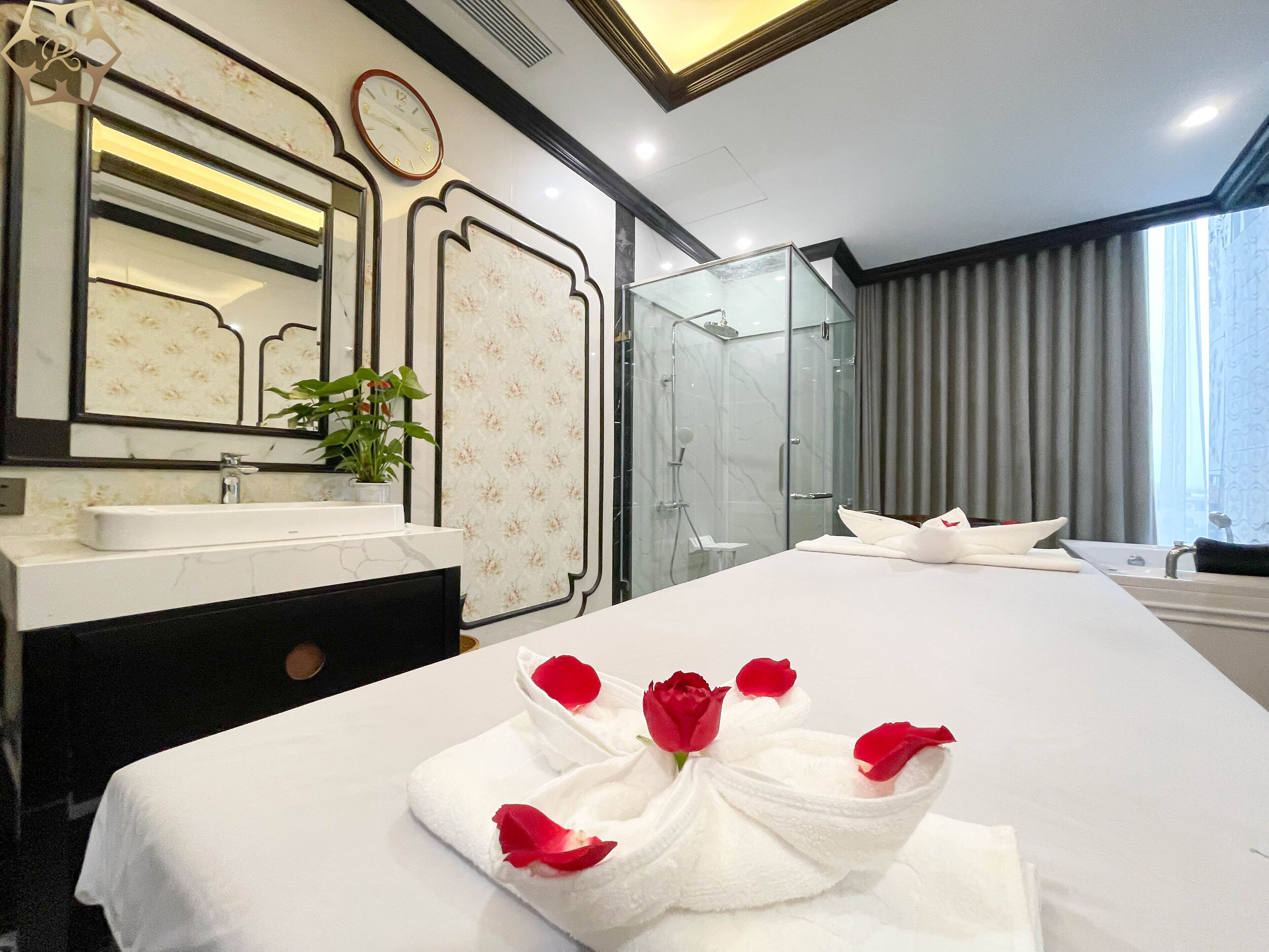 Quarto de hotel com uma cama grande, banheiro com chuveiro de vidro, pia e espelho com relógio.