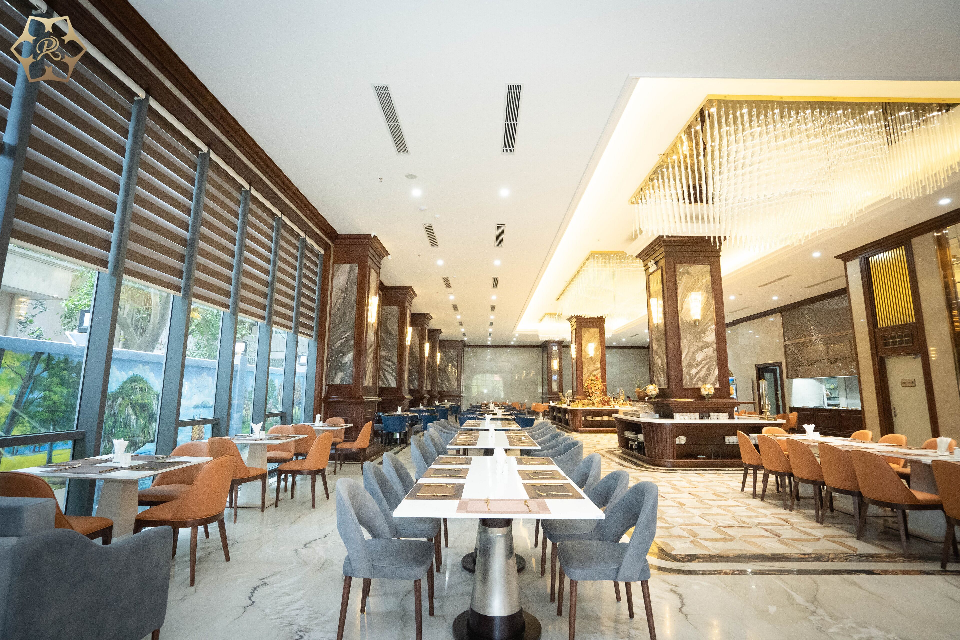 Ampla sala de jantar de hotel, com várias mesas e cadeiras, janelas grandes e um candelabro.