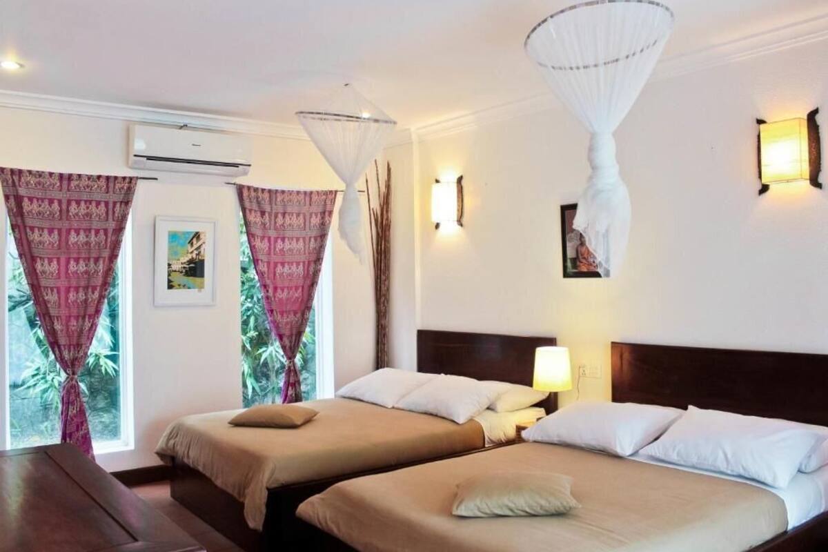 Quarto de hotel com duas camas, um banco de madeira, uma rede mosquiteira e um ar condicionado fixado na parede.