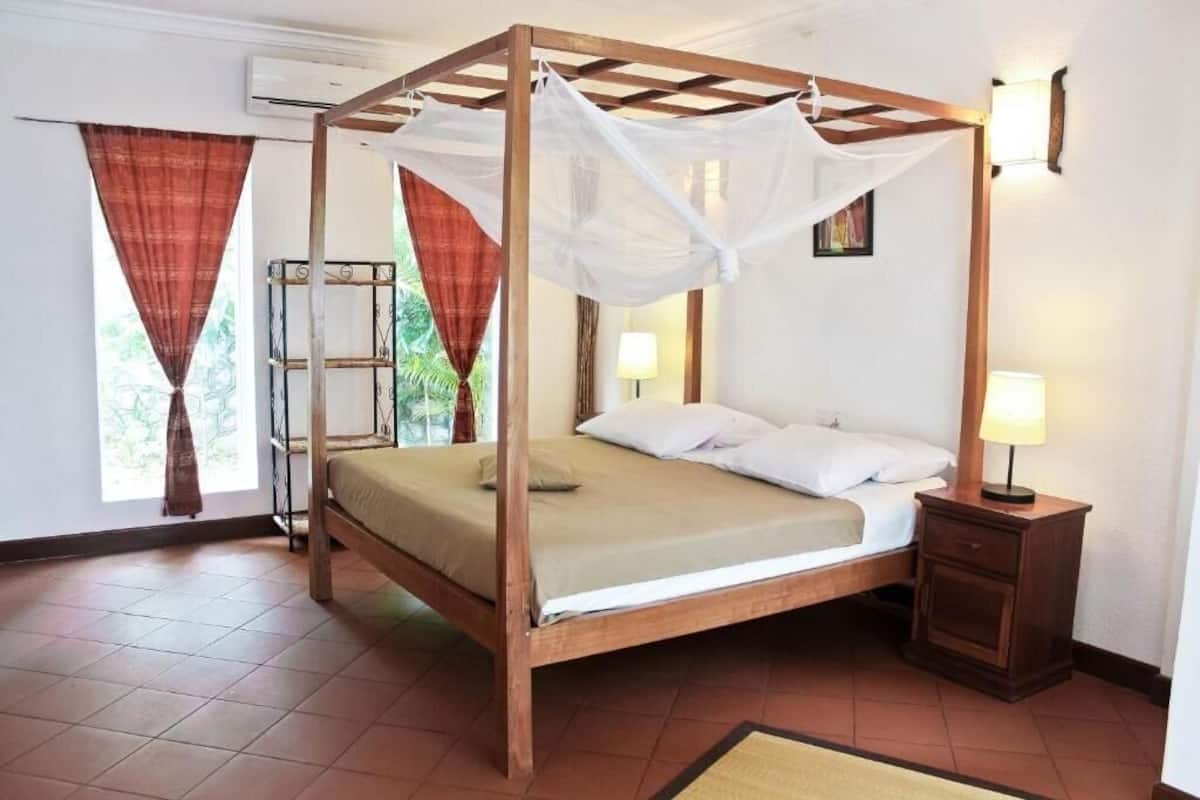 Uma cama com dossel e cortinas, uma mesa de cabeceira com abajur e uma janela com cortinas.