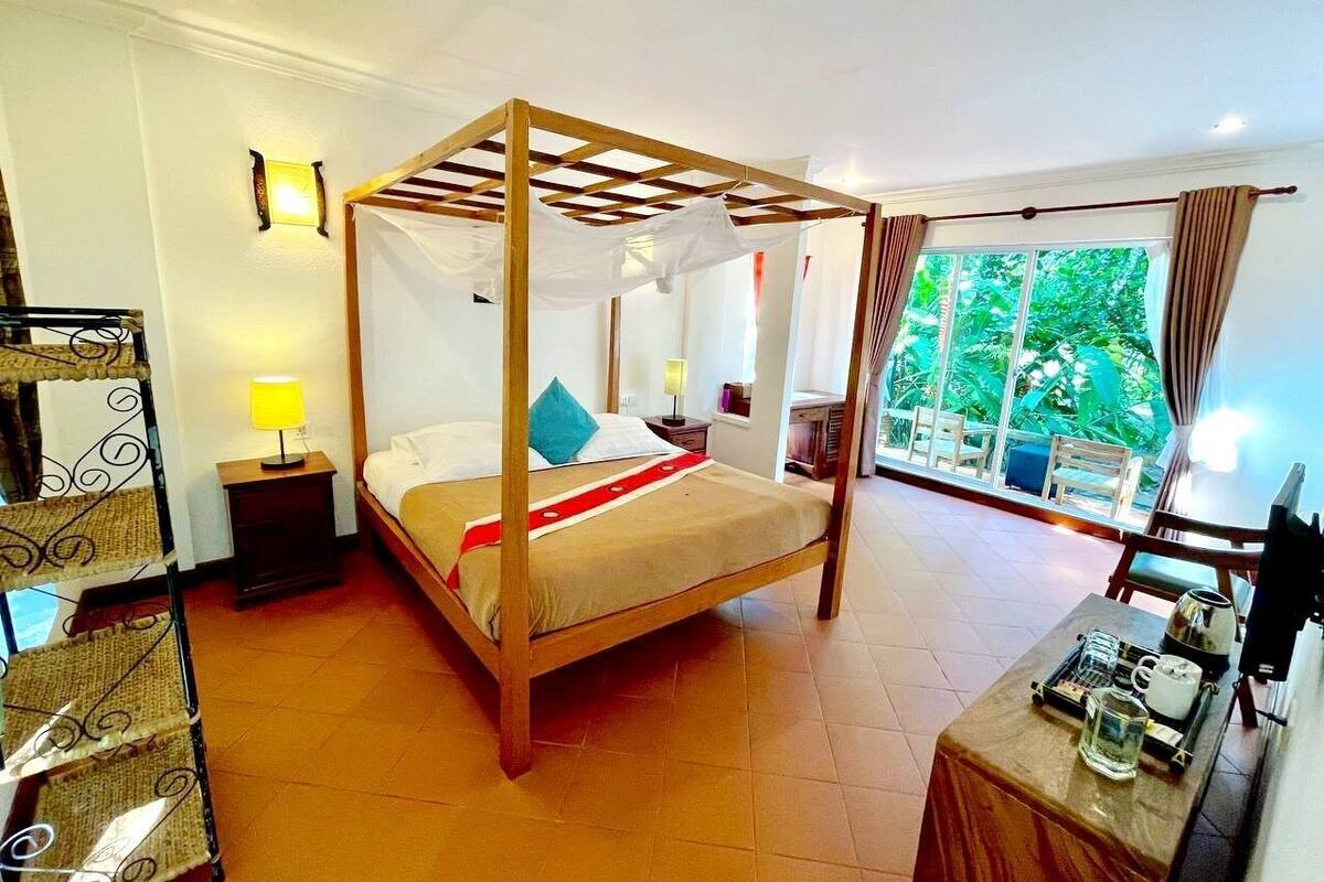 Uma cama com dossel, um tapete vermelho, uma mesa de cabeceira com abajur e vista para o verde pela janela.