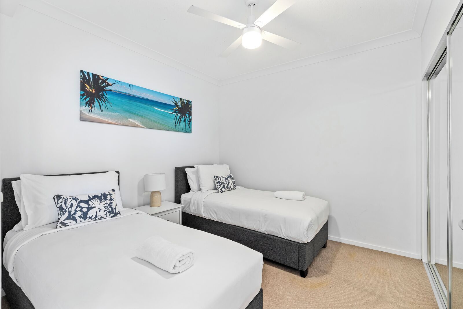 Quarto de hotel com duas camas, um ventilador de teto, um quadro de uma praia e uma janela com vista.