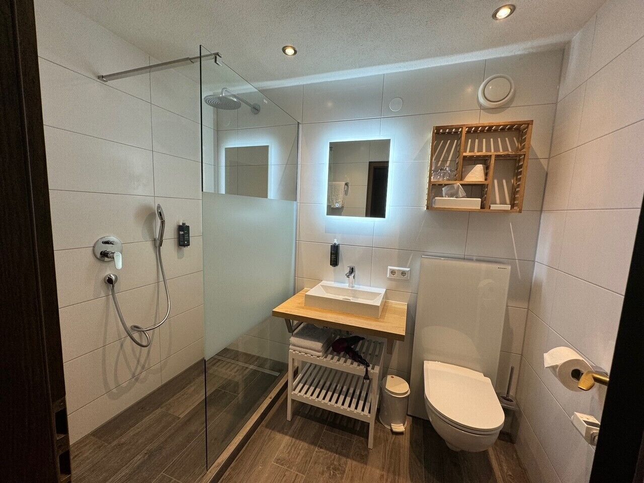Um banheiro moderno com chuveiro em box de vidro, pia com bancada de madeira e vaso sanitário.