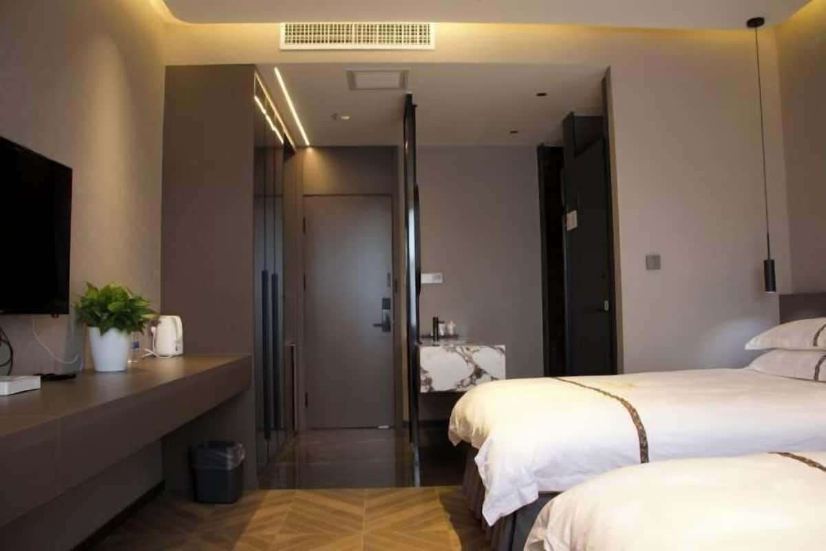 Quarto de hotel moderno com uma cama grande, uma mesa de cabeceira com uma planta, uma TV de tela plana e um banheiro visível através de uma porta de vidro.