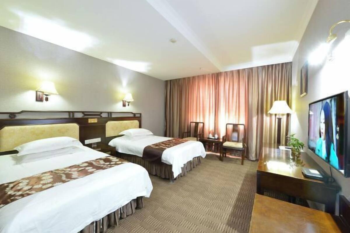 Quarto de hotel com duas camas, uma escrivaninha, televisão e uma janela grande com cortinas.