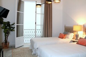 Quarto de hotel com duas camas, uma TV fixada na parede, uma varanda com guarda-corpo de metal e um abajur sobre uma mesinha de cabeceira.