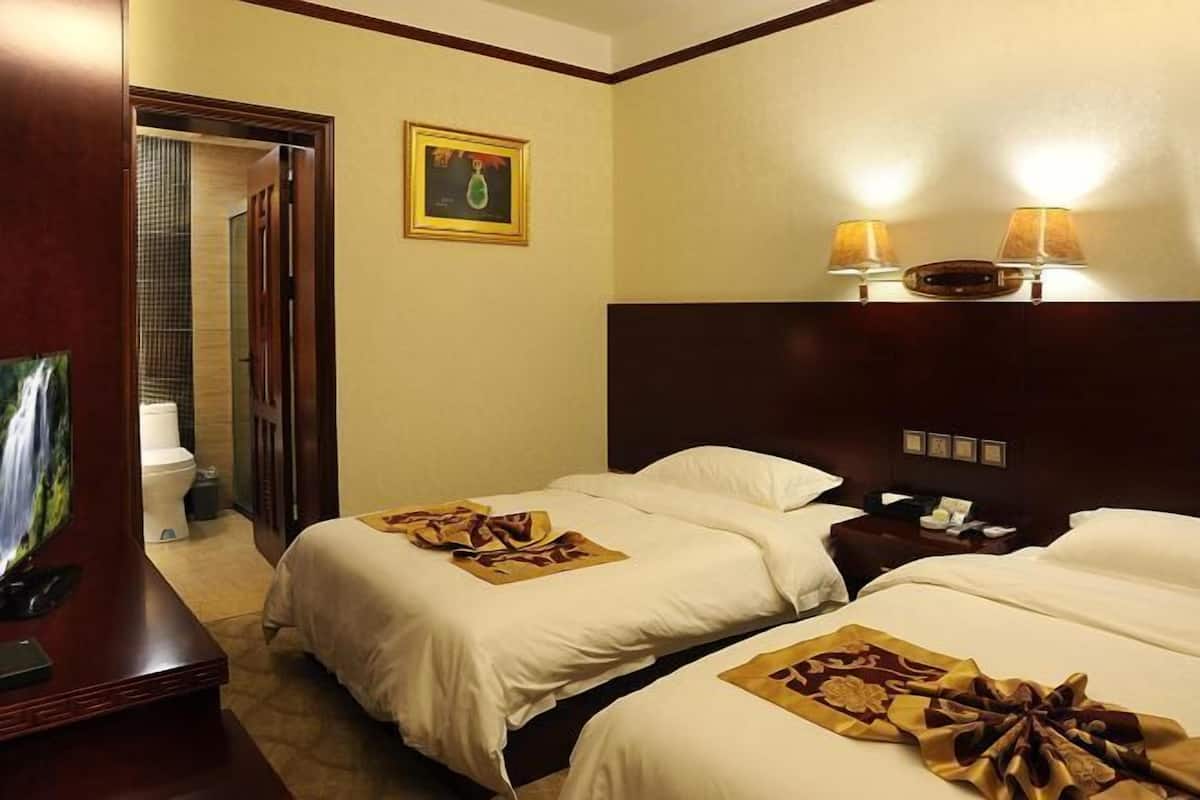 Quarto de hotel com duas camas, cabeceira de madeira, mesa de cabeceira, quadro na parede e televisão sobre suporte.
