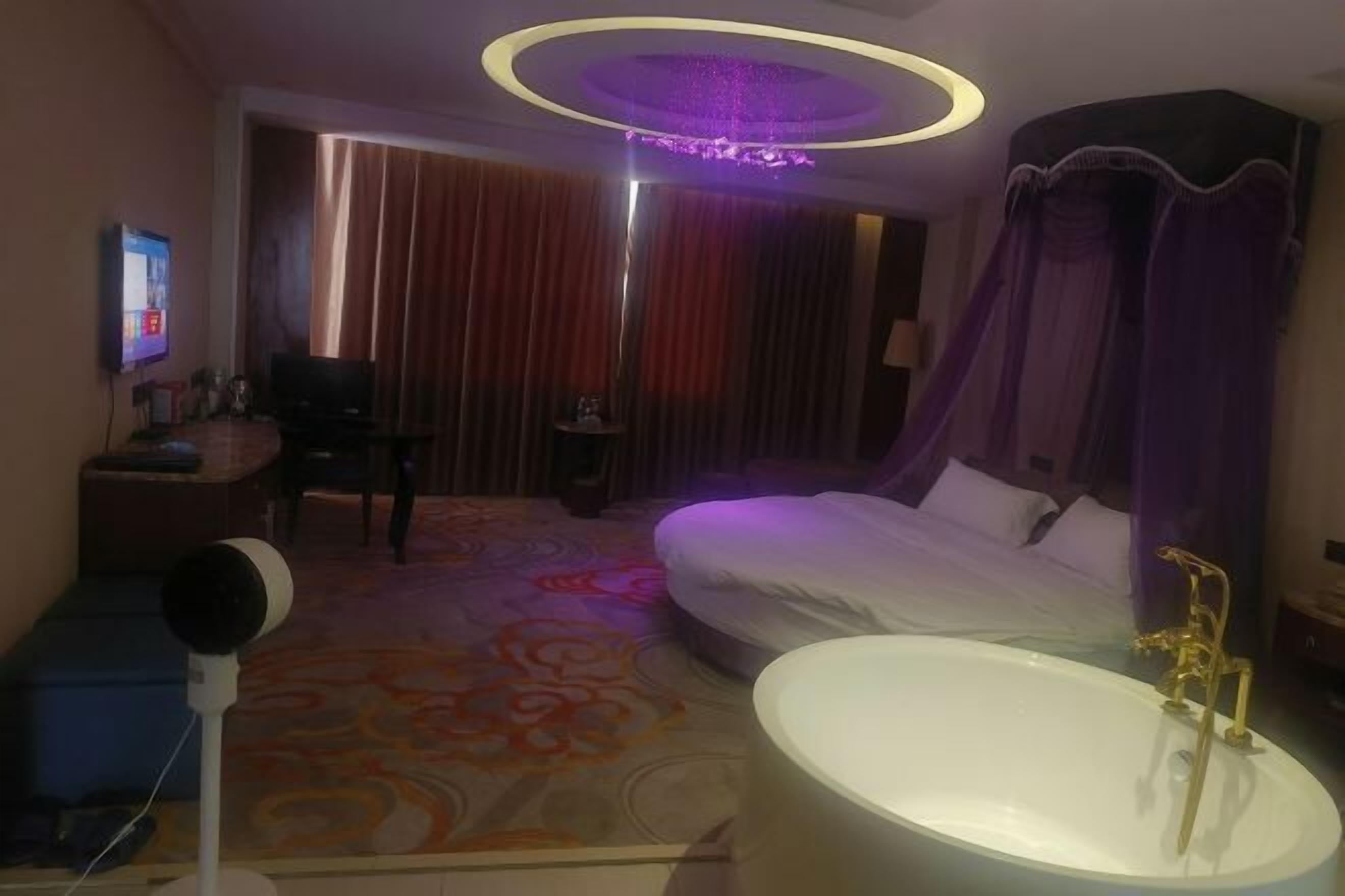 Quarto de hotel com cama, banheiro com banheira, escrivaninha, televisão e cortina.