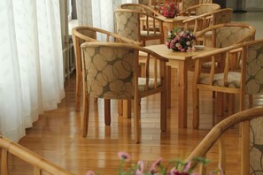 Uma sala de jantar com mesas e cadeiras de madeira, um arranjo floral e uma escadaria que leva a um andar superior.