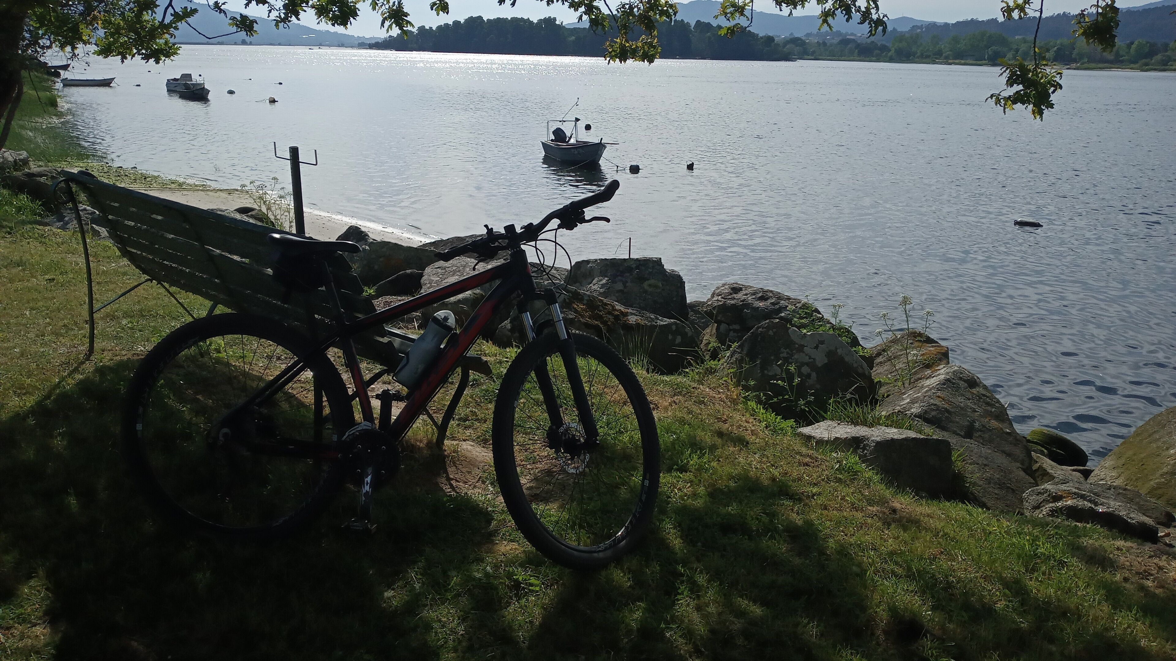 Uma bicicleta estacionada em área gramada à beira do lago, com um banco e pedras por perto.
