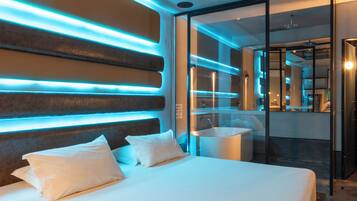 Quarto de hotel moderno com uma cama grande, divisória de vidro e iluminação ambiente.