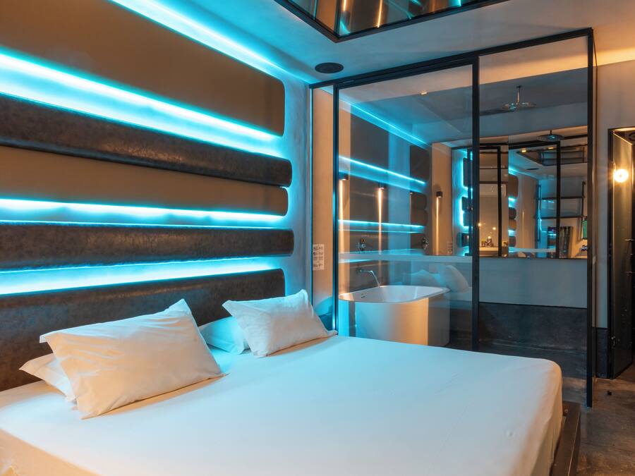 Quarto de hotel moderno com uma cama grande, divisória de vidro e iluminação ambiente.