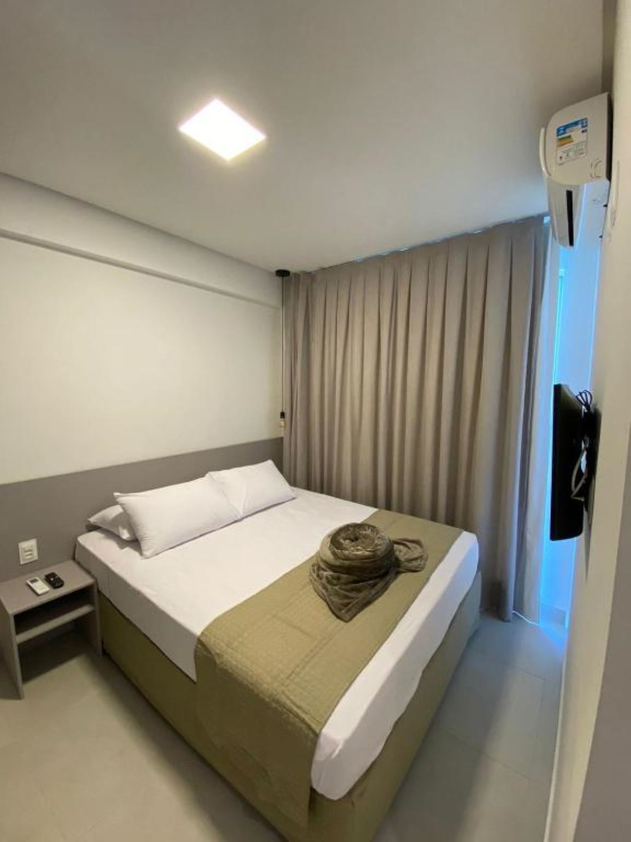 Quarto de hotel com cama, mesa de cabeceira, ar condicionado na parede e janela com cortinas.